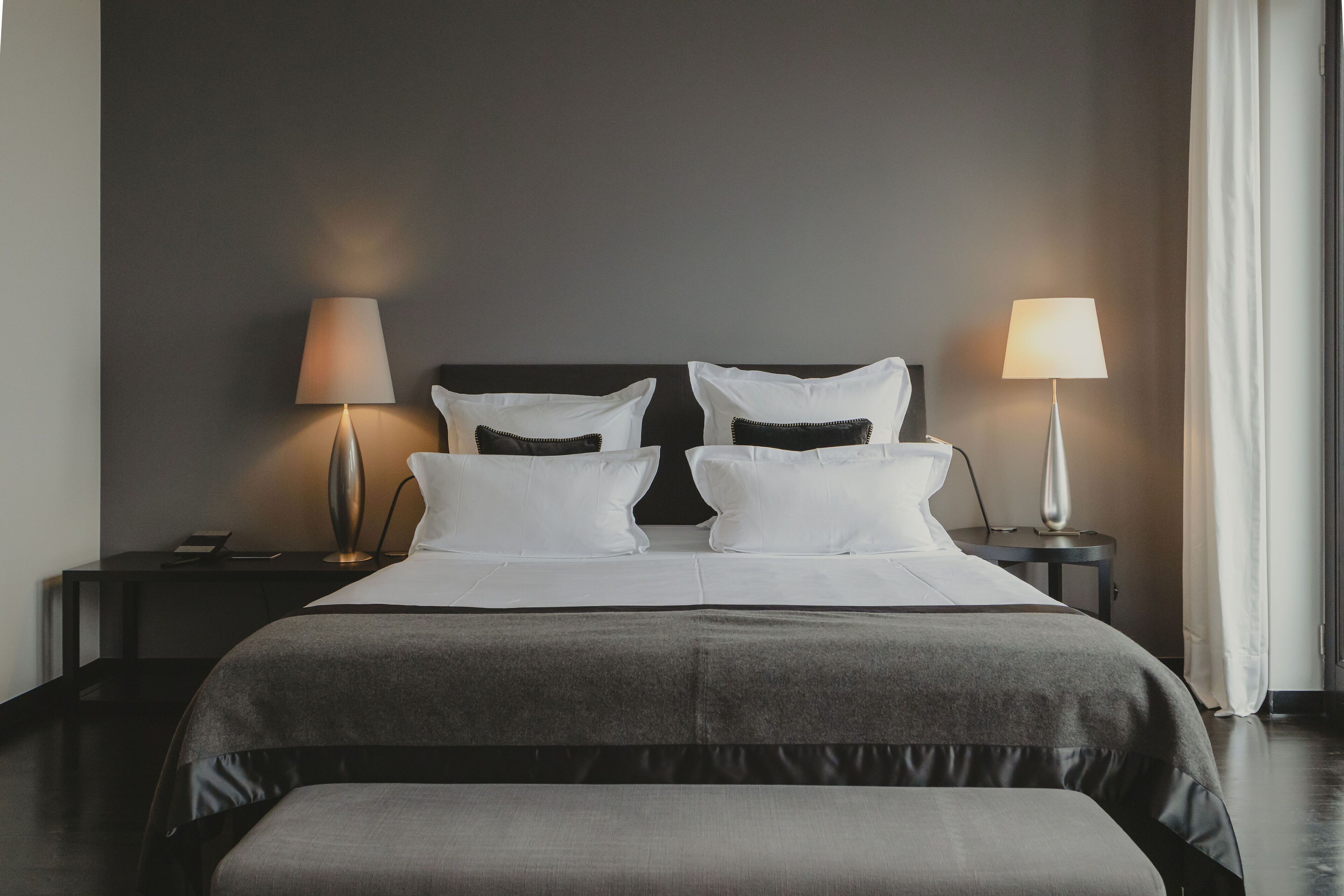 Cama bem arrumada com colcha cinza e almofadas brancas, com duas mesas de cabeceira com luminárias, em um quarto de hotel moderno.