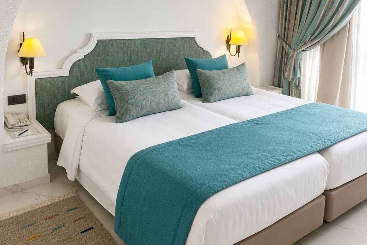 Uma cama de casal com cabeceira branca e duas luminárias de mesa, um telefone e um tapete estampado.