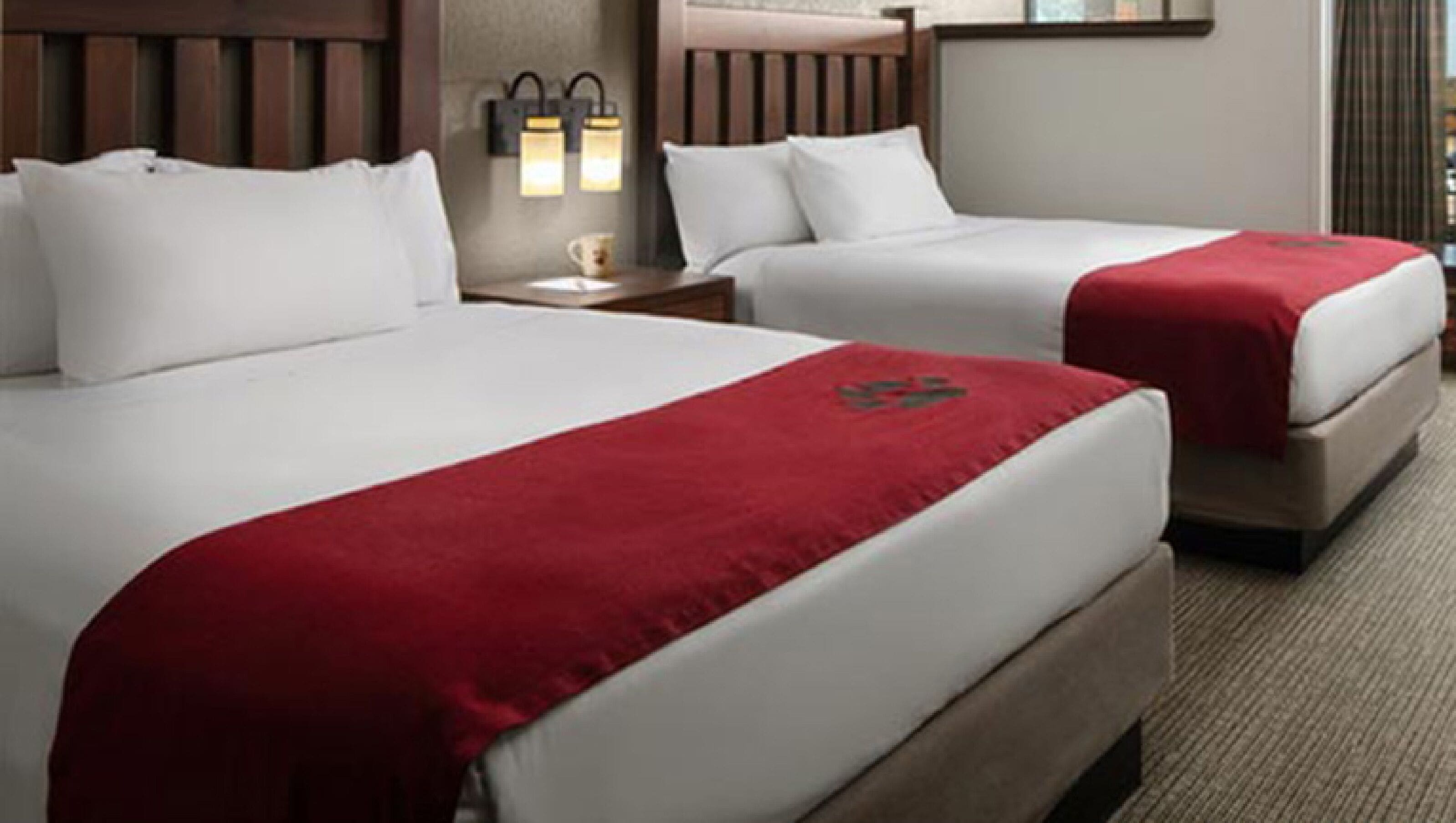 Quarto de hotel com duas camas, cada uma com roupa de cama branca e manta vermelha. Há uma mesa de cabeceira com abajur entre as camas.