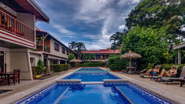 Uma piscina com deck revestido, rodeada por espreguiçadeiras e guarda-sóis, com edifícios e árvores ao fundo.