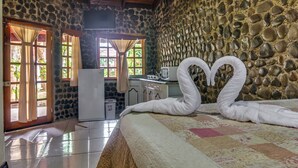 Quarto com parede de pedra, cama com colcha, geladeira e janela com cortinas.