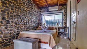Quarto com parede de pedra, cama com cabeceira decorativa, um banco e janela com vista.