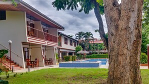 Um edifício de dois andares com piscina, área de estar externa e uma grande árvore em primeiro plano.
