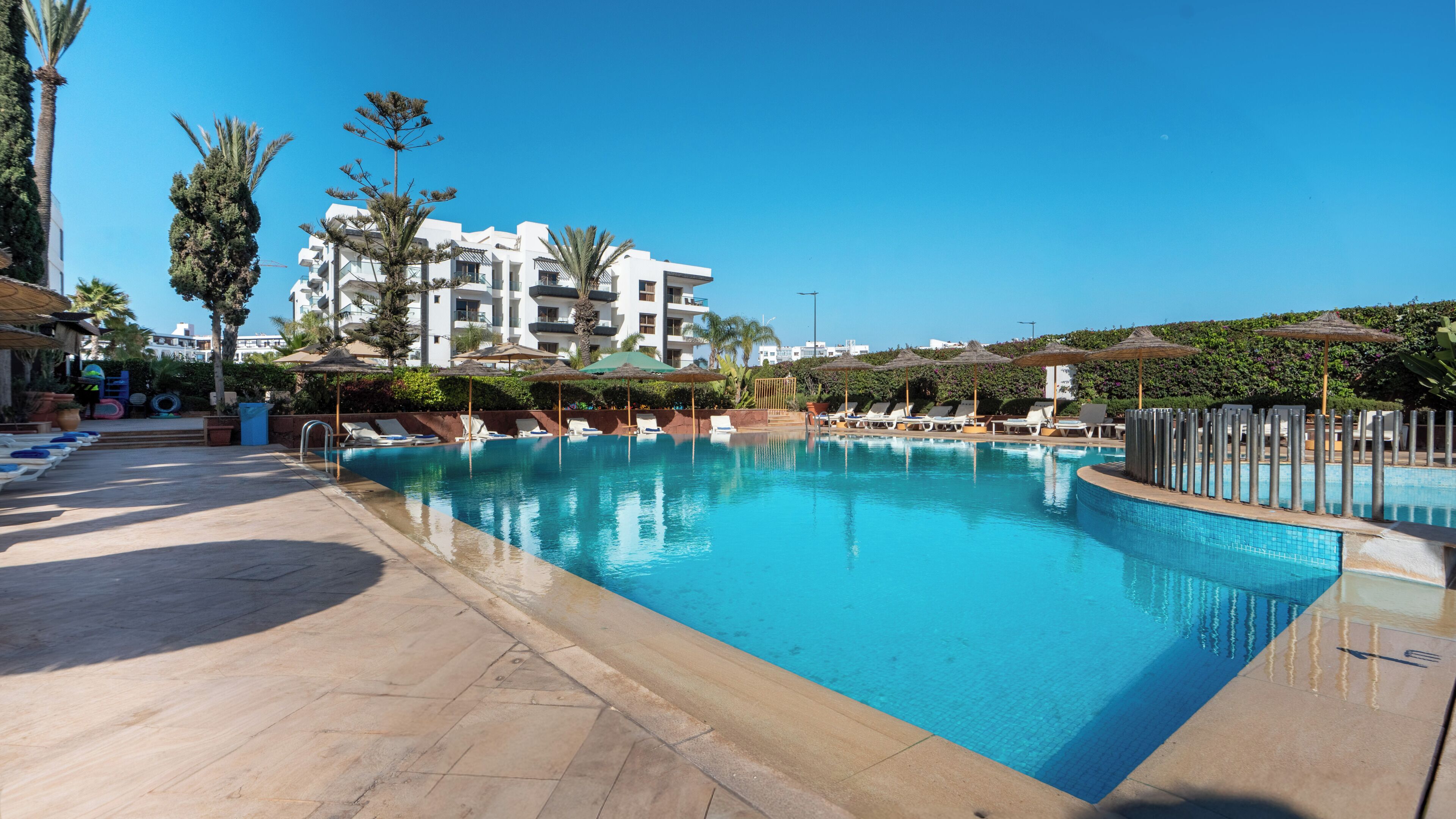 Piscina azul límpida com espreguiçadeiras e guarda-sóis, cercada por um edifício com varandas.