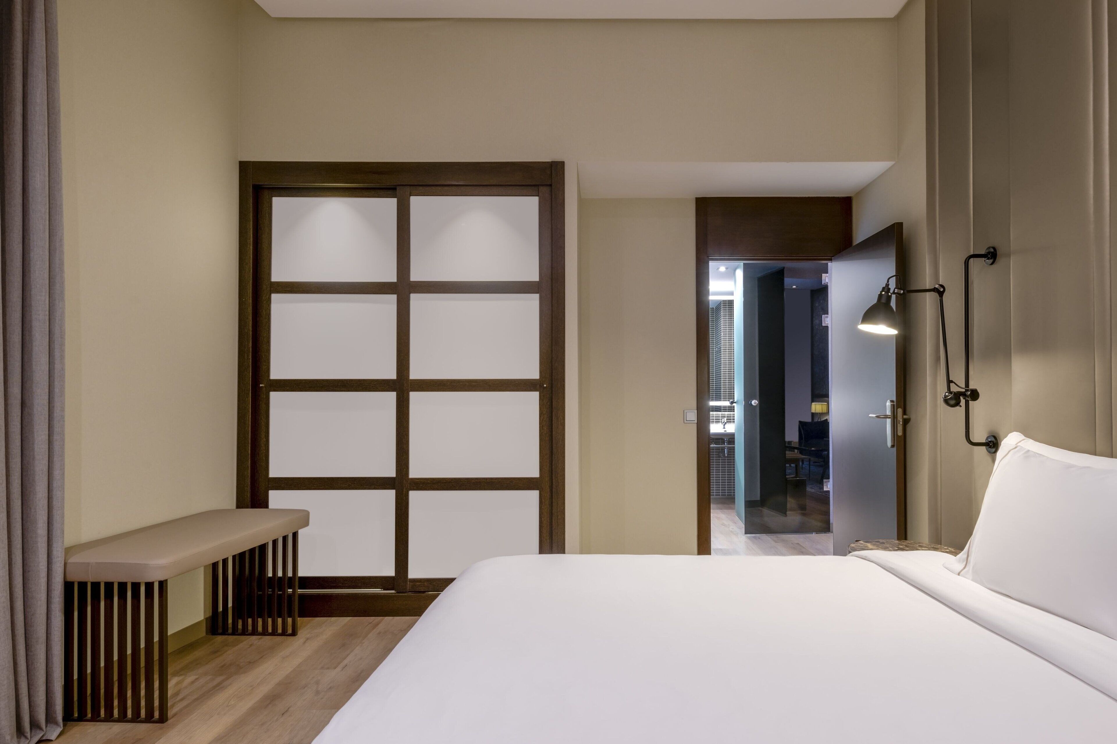 Quarto de hotel com cama, um banco, porta de madeira e luminária fixada na parede.