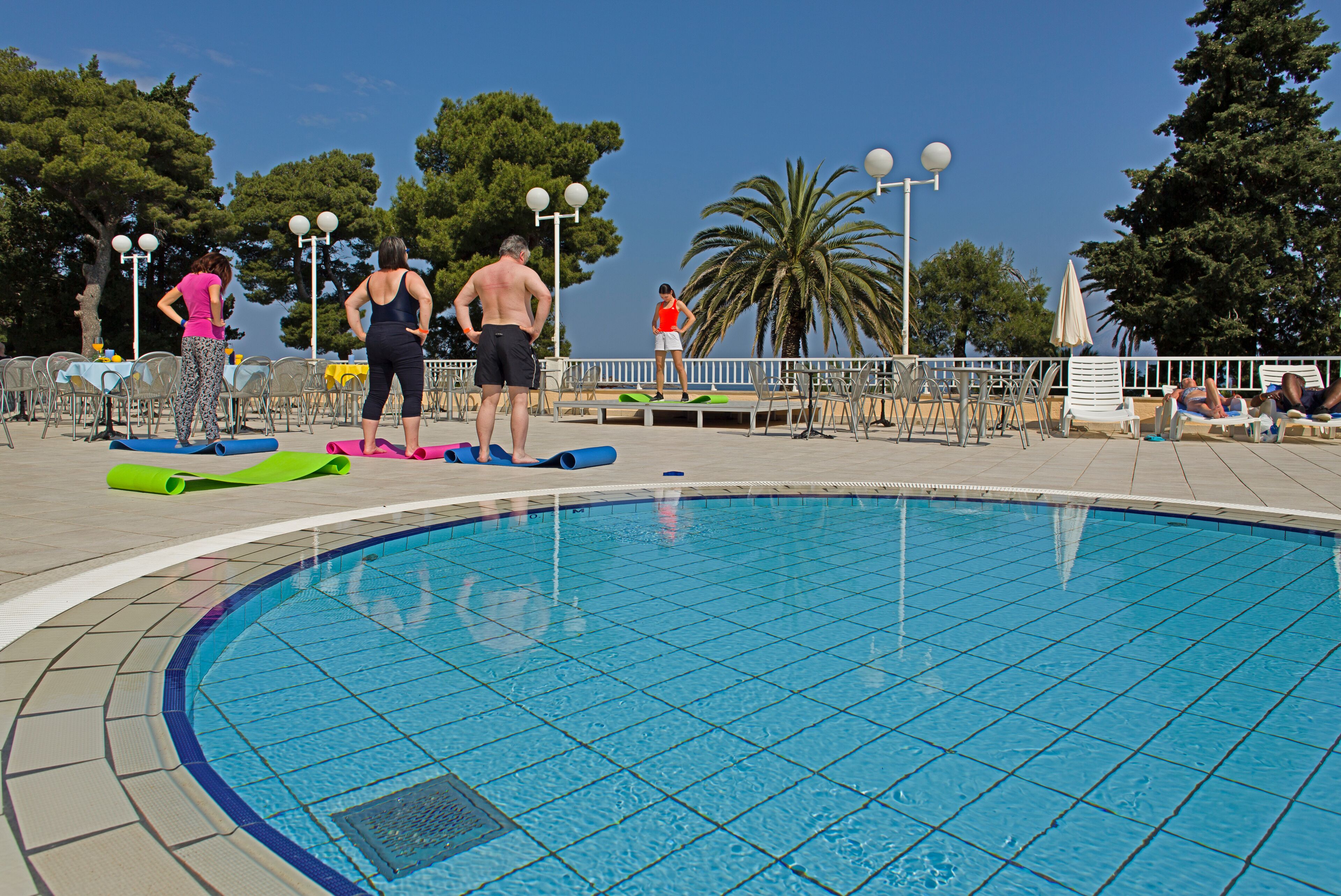 Piscina com pessoas ao redor, algumas em pé e outras sentadas.