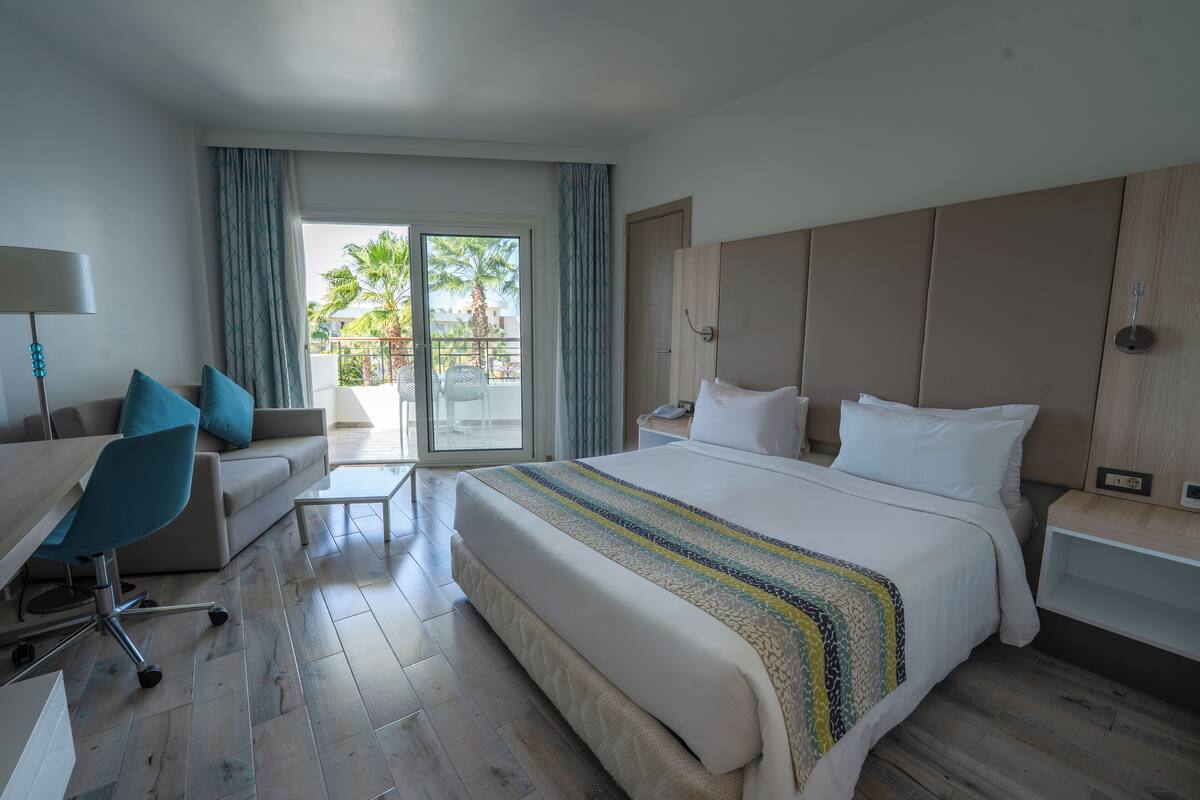Quarto de hotel com uma cama grande, uma escrivaninha com cadeira, um sofá com almofadas azuis, uma mesa de cabeceira e vista para palmeiras através de uma porta de correr.