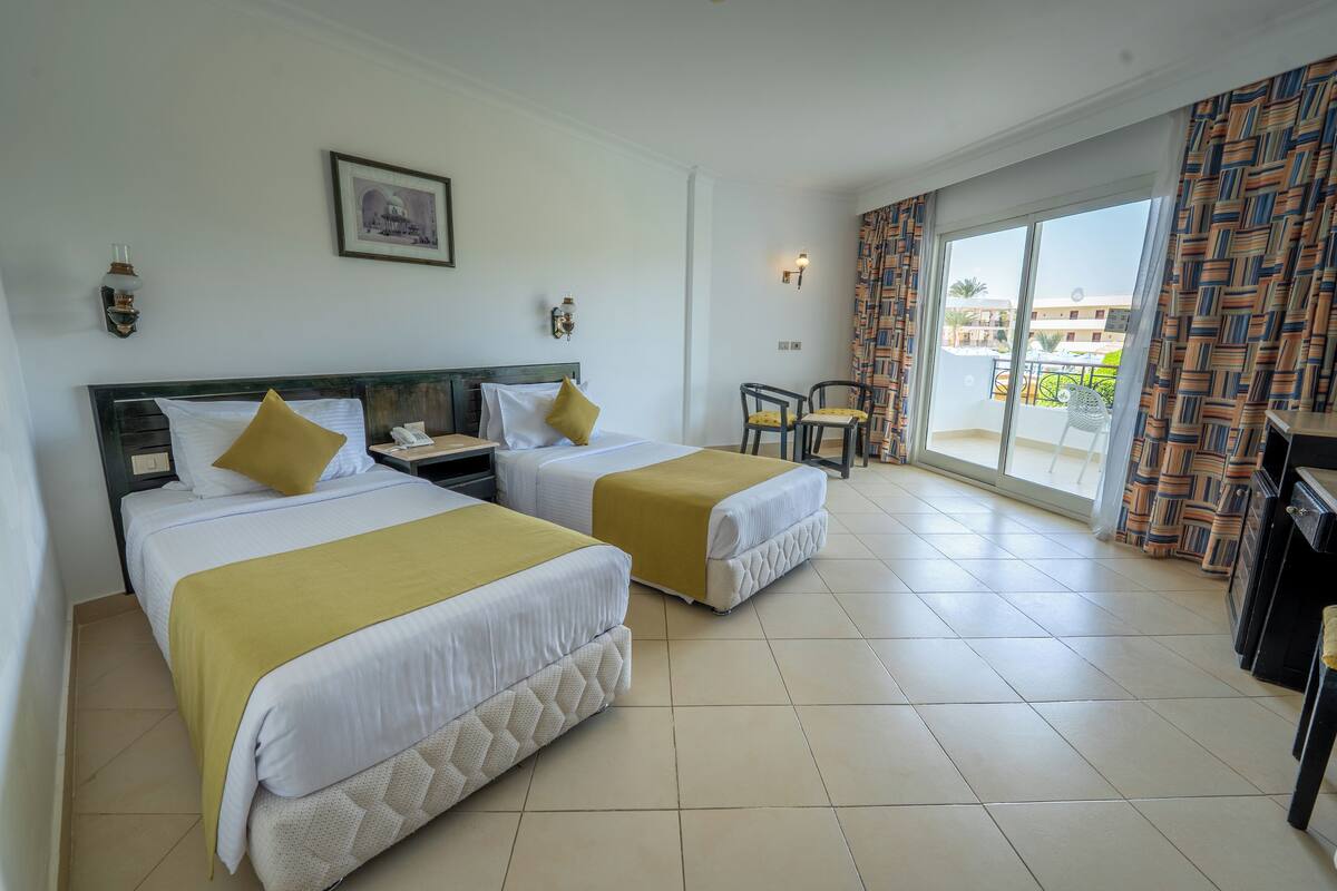 Quarto de hotel com duas camas, uma escrivaninha, uma cadeira e varanda com cortinas estampadas.