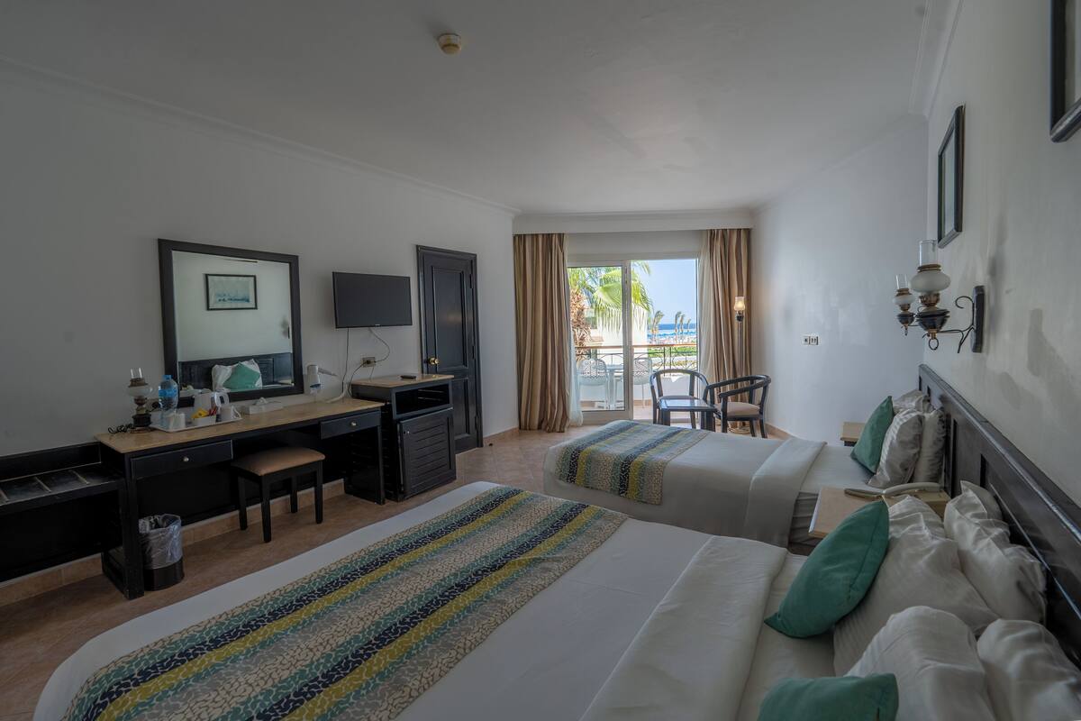 Quarto de hotel com duas camas, uma escrivaninha, uma cadeira, uma televisão e varanda vista para o mar.
