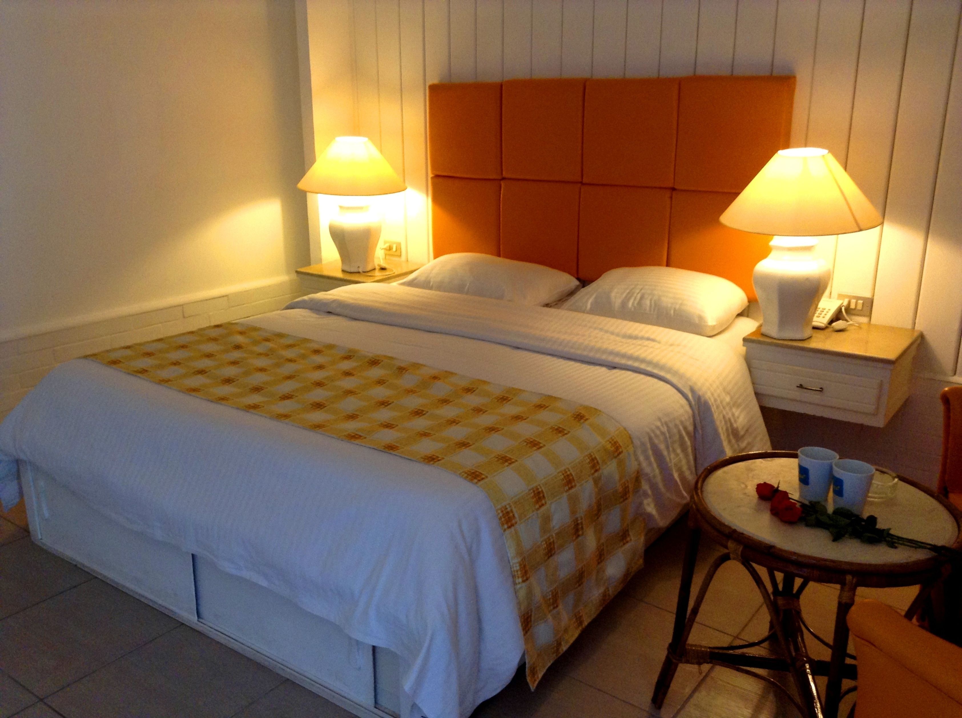 Uma cama bem arrumada com um espalha de retalhos xadrez, duas luminárias de cabeceira, uma mesinha redonda com xícaras e uma rosa, e uma cadeira de madeira.