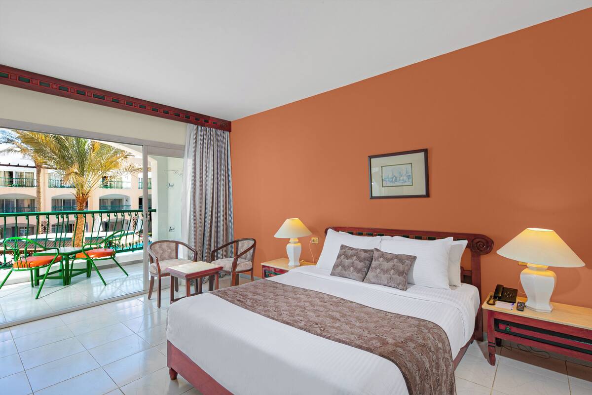 Quarto de hotel com uma cama, duas mesas de cabeceira com luminárias, uma cadeira e uma varanda com vista para palmeiras e edifícios.