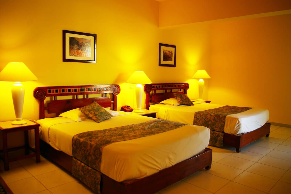 Quarto de hotel com duas camas, cada uma com cabeceira de madeira, roupa de cama branca e luminária na mesa de cabeceira. Há quadros nas paredes e piso de azulejos.