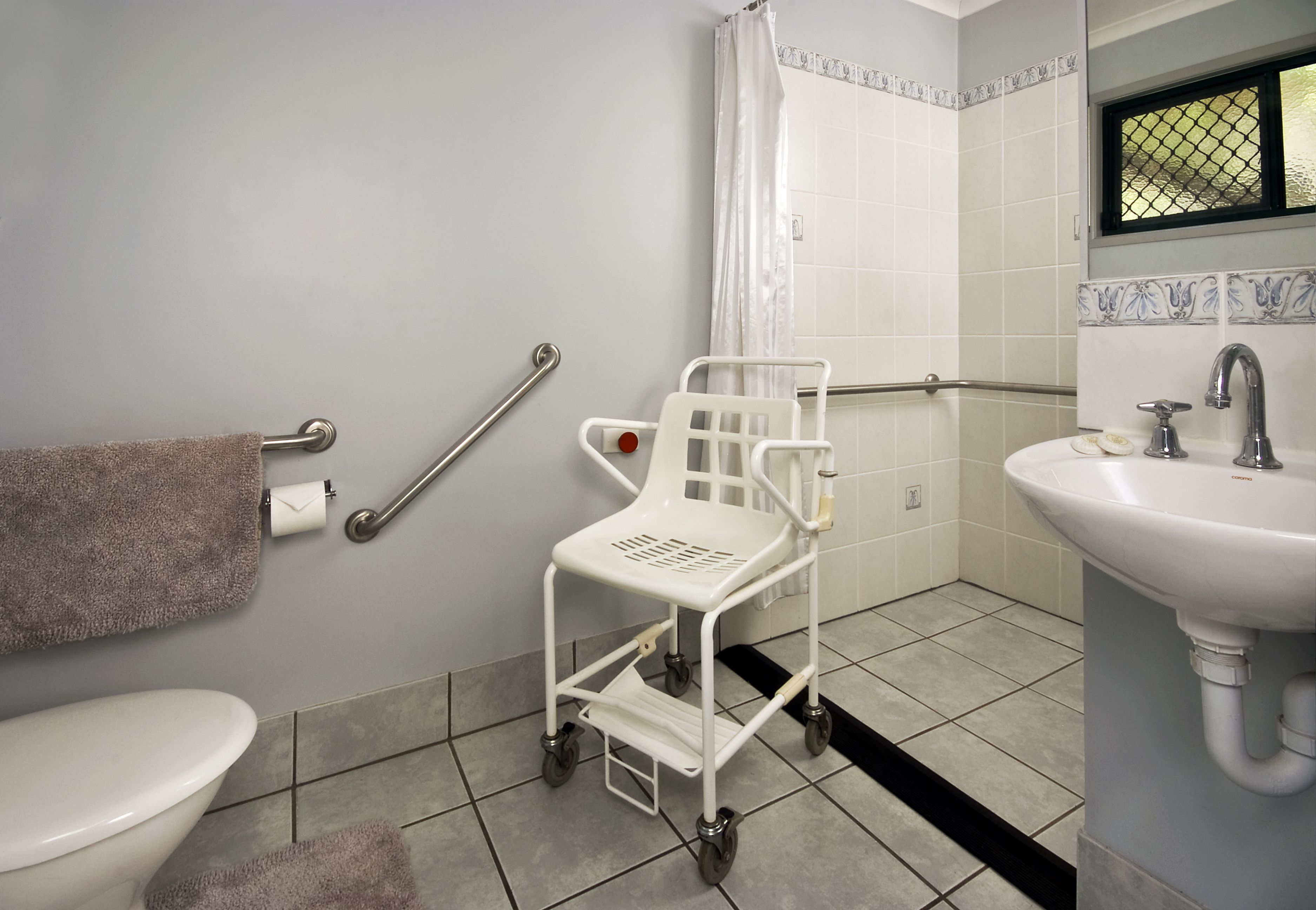 Uma casa de banho com sanita, lavatório, chuveiro com cortina, um auxílio de mobilidade branco e barra de apoio.