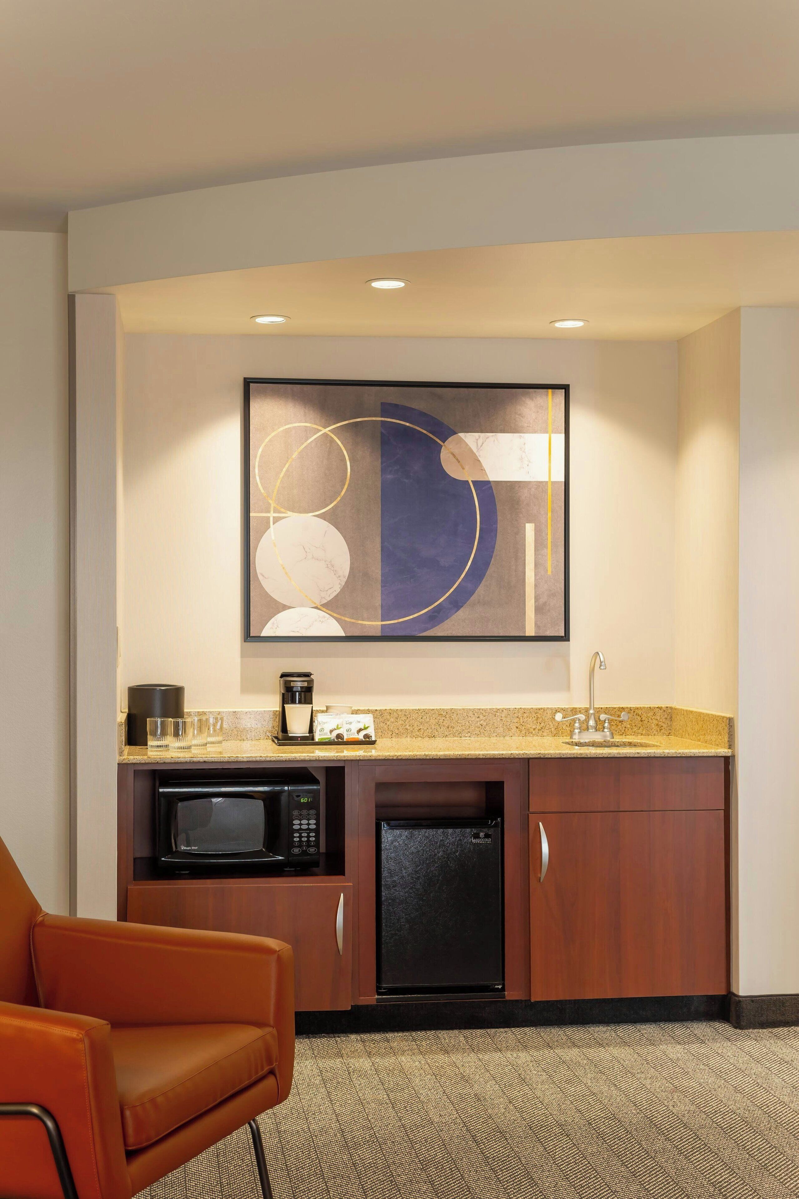Quarto de hotel moderno com cozinha compacta, incluindo micro-ondas, cafeteira e frigorífico. Há uma obra de arte abstrata emoldurada sobre o balcão e uma cadeira laranja individual no quarto.
