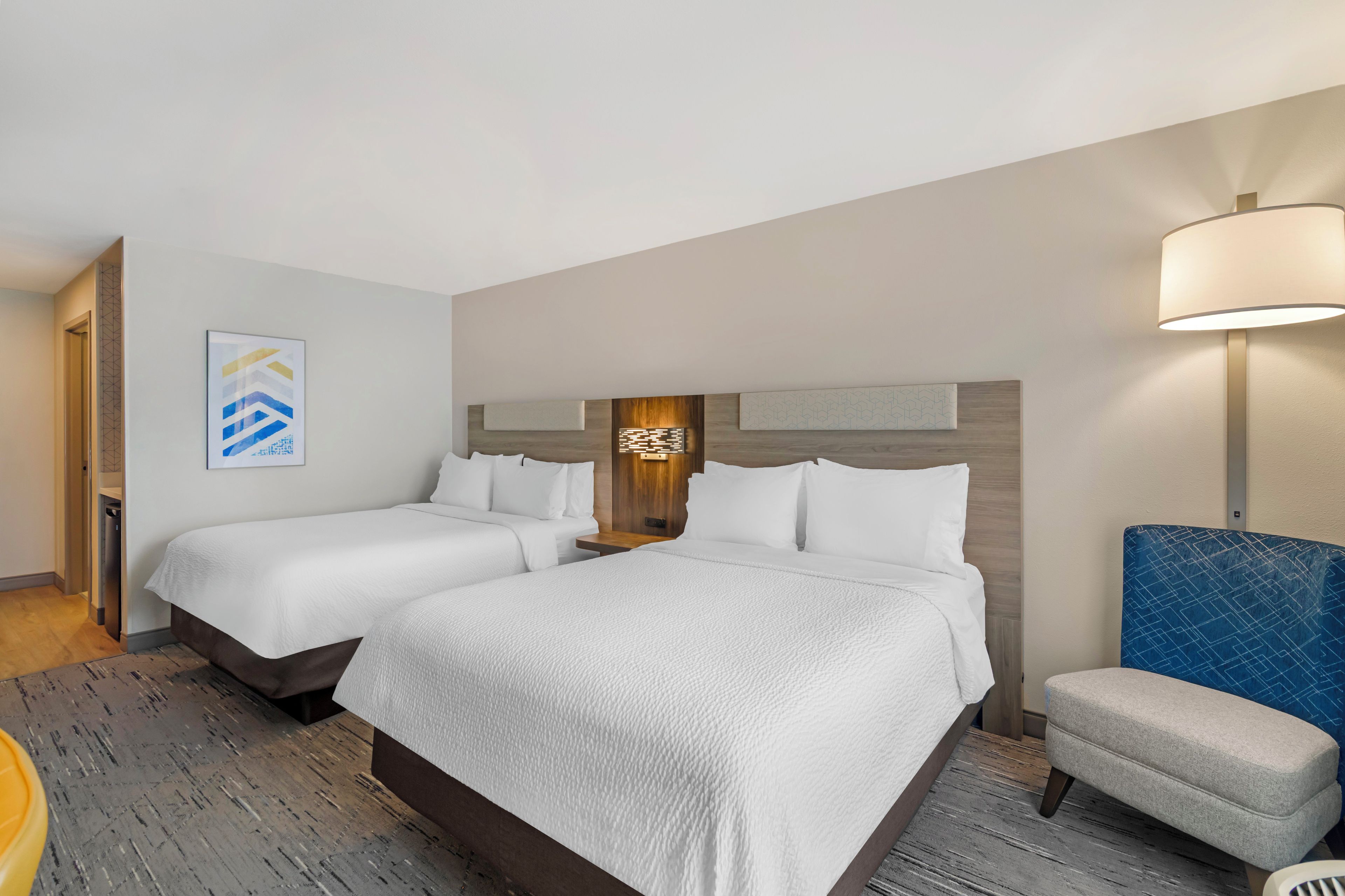 Quarto de hotel com duas camas, uma cadeira, um abajur e uma obra de arte na parede.