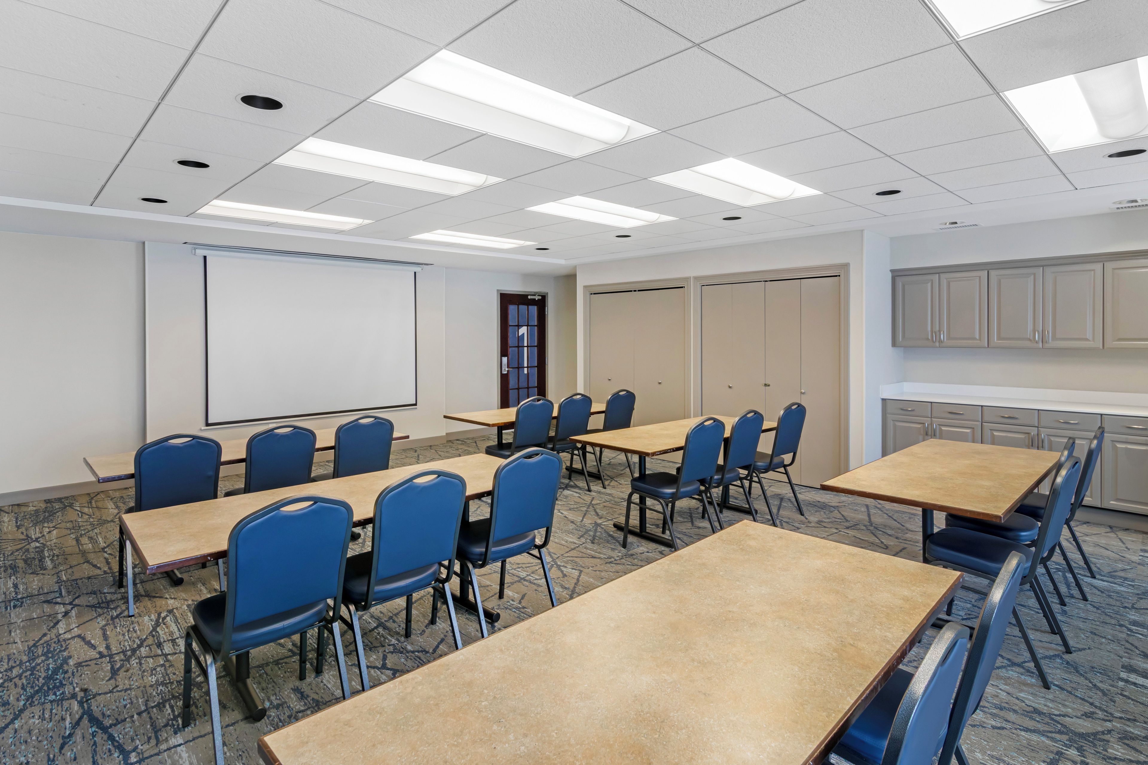 Sala de conferência com mesas, cadeiras, tela de projeção e um espaço com cozinha.