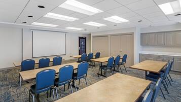 Sala de conferência com mesas, cadeiras, tela de projeção e um espaço com cozinha.