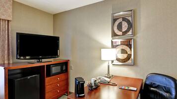 Quarto de hotel com televisão de tela plana, mini-geladeira, cafeteira, escrivaninha com luminária, telefone e uma cadeira.