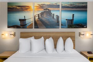 Quarto de hotel com cabeceira de madeira, cama com roupa de cama branca e pintura na parede retratando um cais ao pôr do sol.