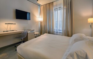 Quarto de hotel com cama, escrivaninha, cadeira, televisão de tela plana, abajur e janela com cortinas.