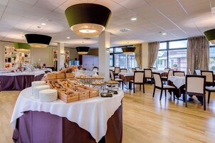 Uma ampla sala de jantar com várias mesas e cadeiras, um buffet com diversos alimentos, e janelas grandes que proporcionam luz natural.