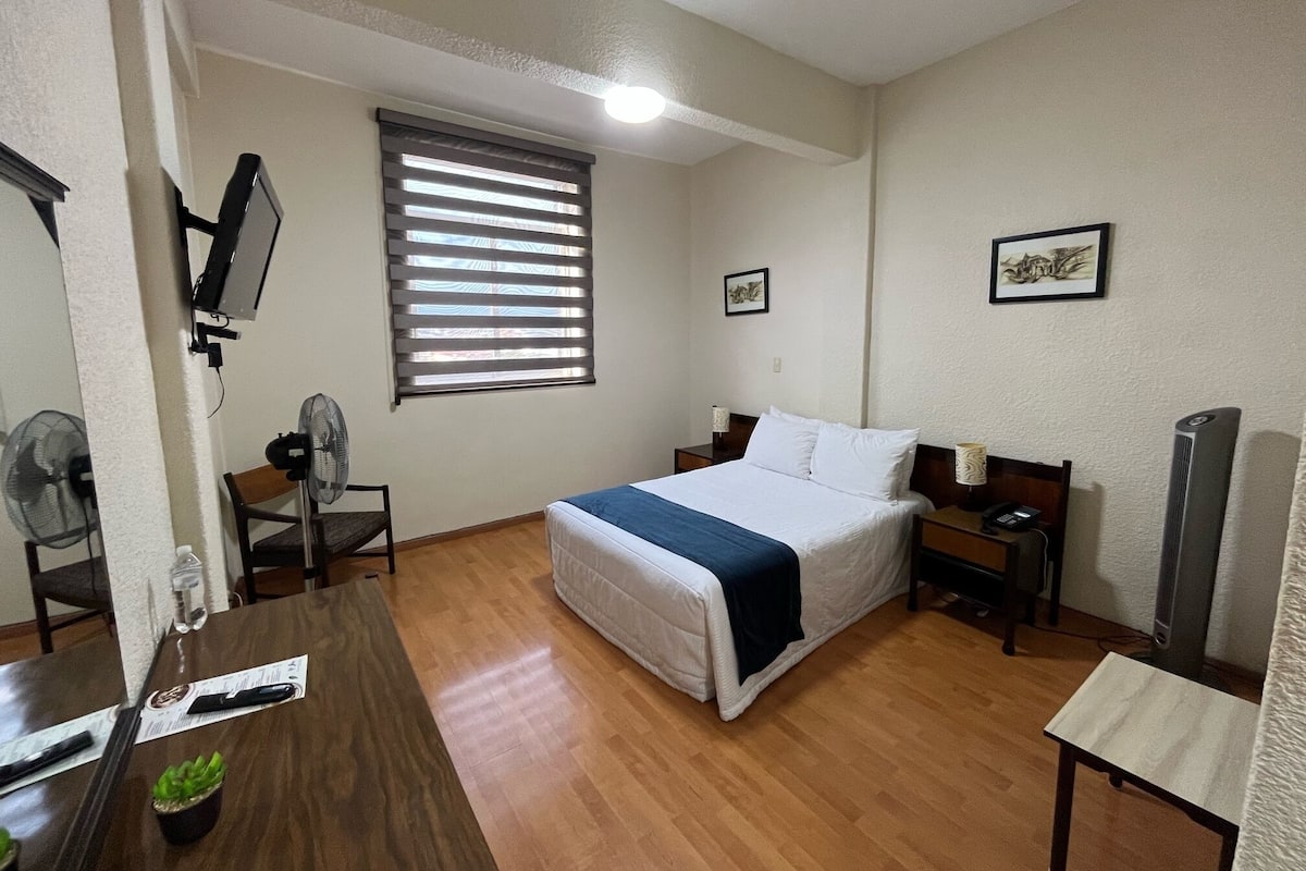 Quarto de hotel com cama, mesa de cabeceira, abajur e janela com persianas.