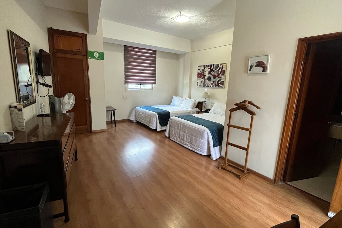 Quarto de hotel com duas camas, uma escrivaninha, uma cadeira, uma escada de madeira, uma janela com persianas e um quadro na parede.