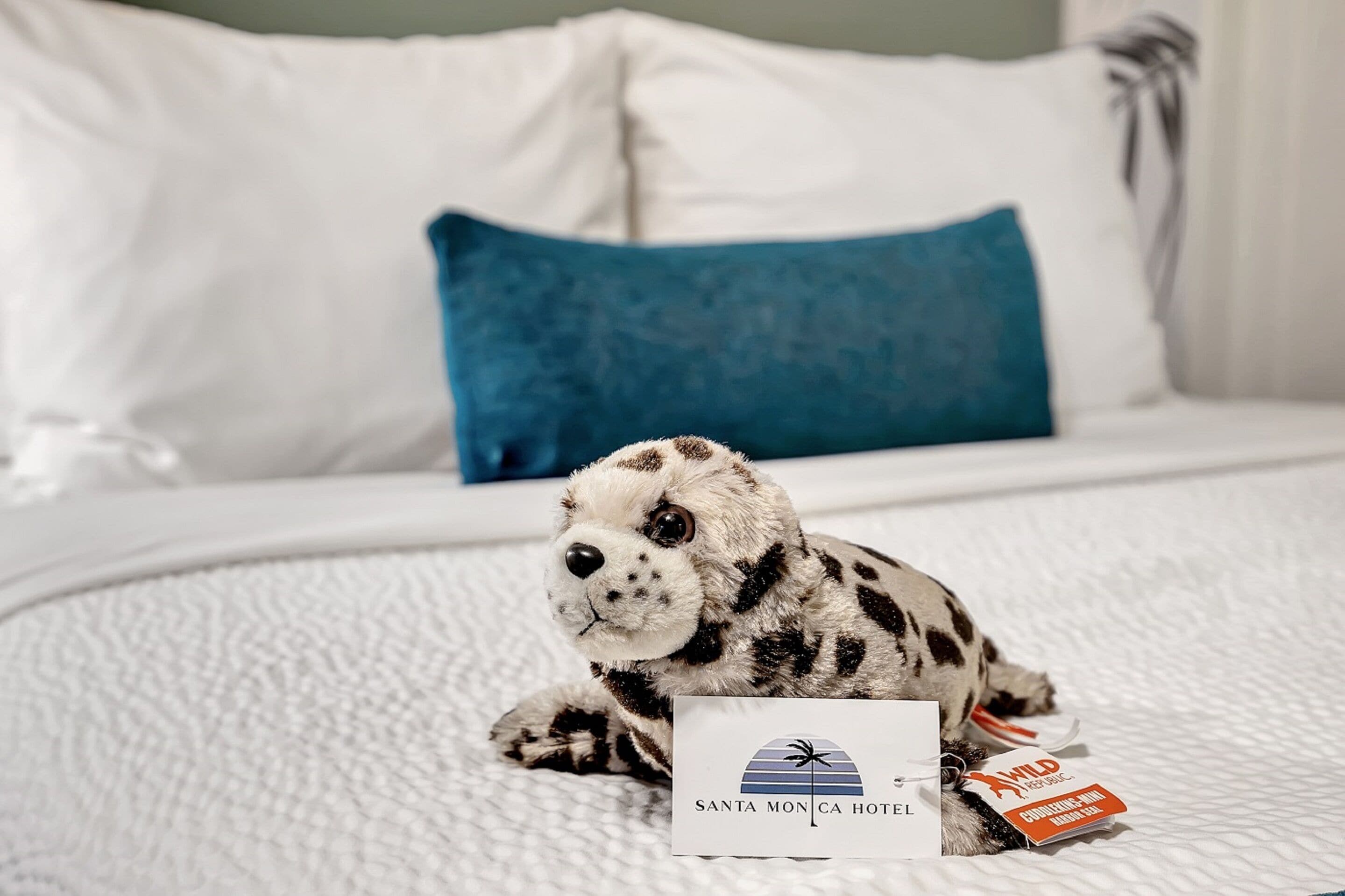 Um brinquedo de pelúcia em uma cama com travesseiro branco e azul, uma etiqueta e o logotipo do Hotel Santa Monica CA.