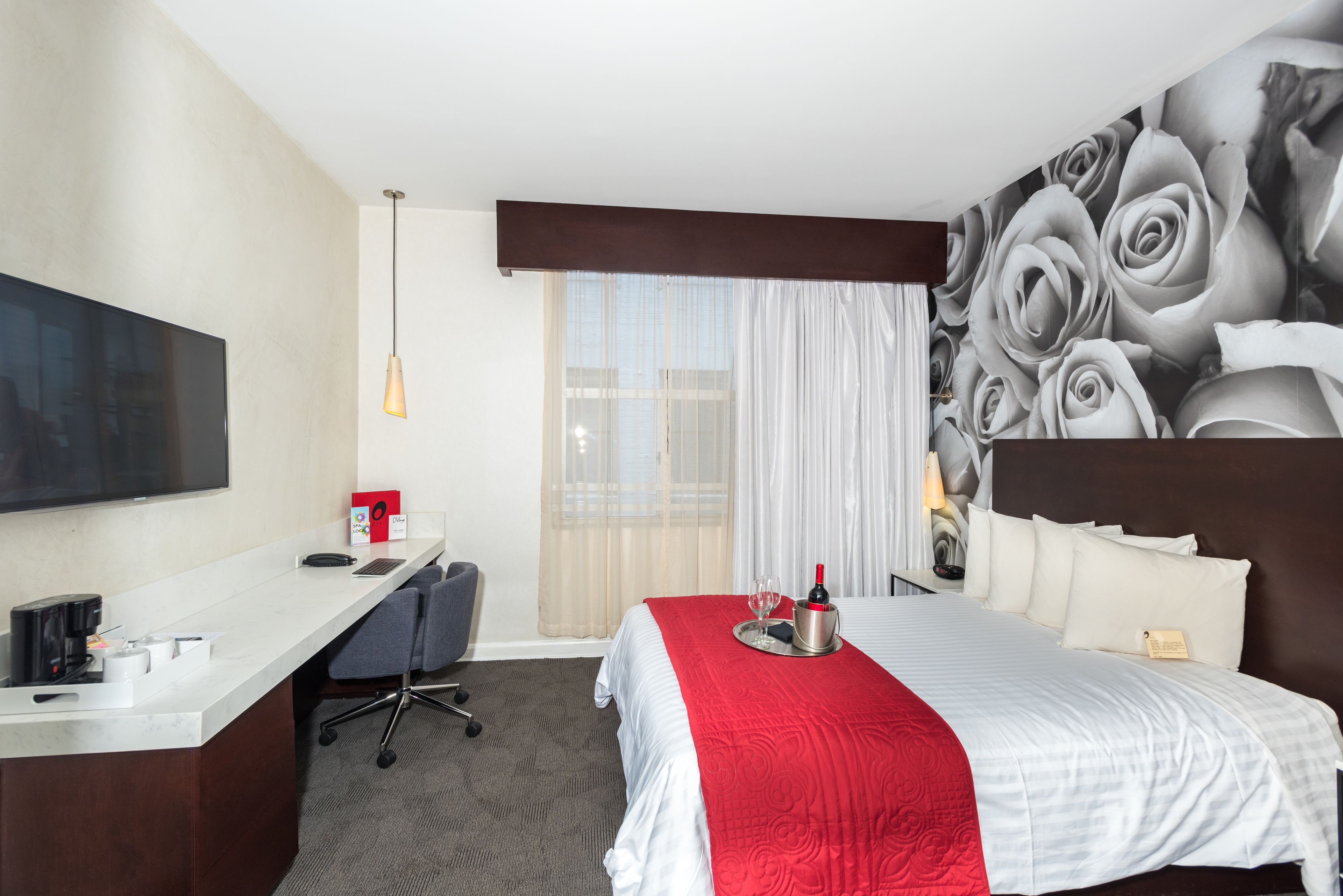 Quarto de hotel com cama, escrivaninha, televisão, janela com cortinas e parede com padrão de rosas.
