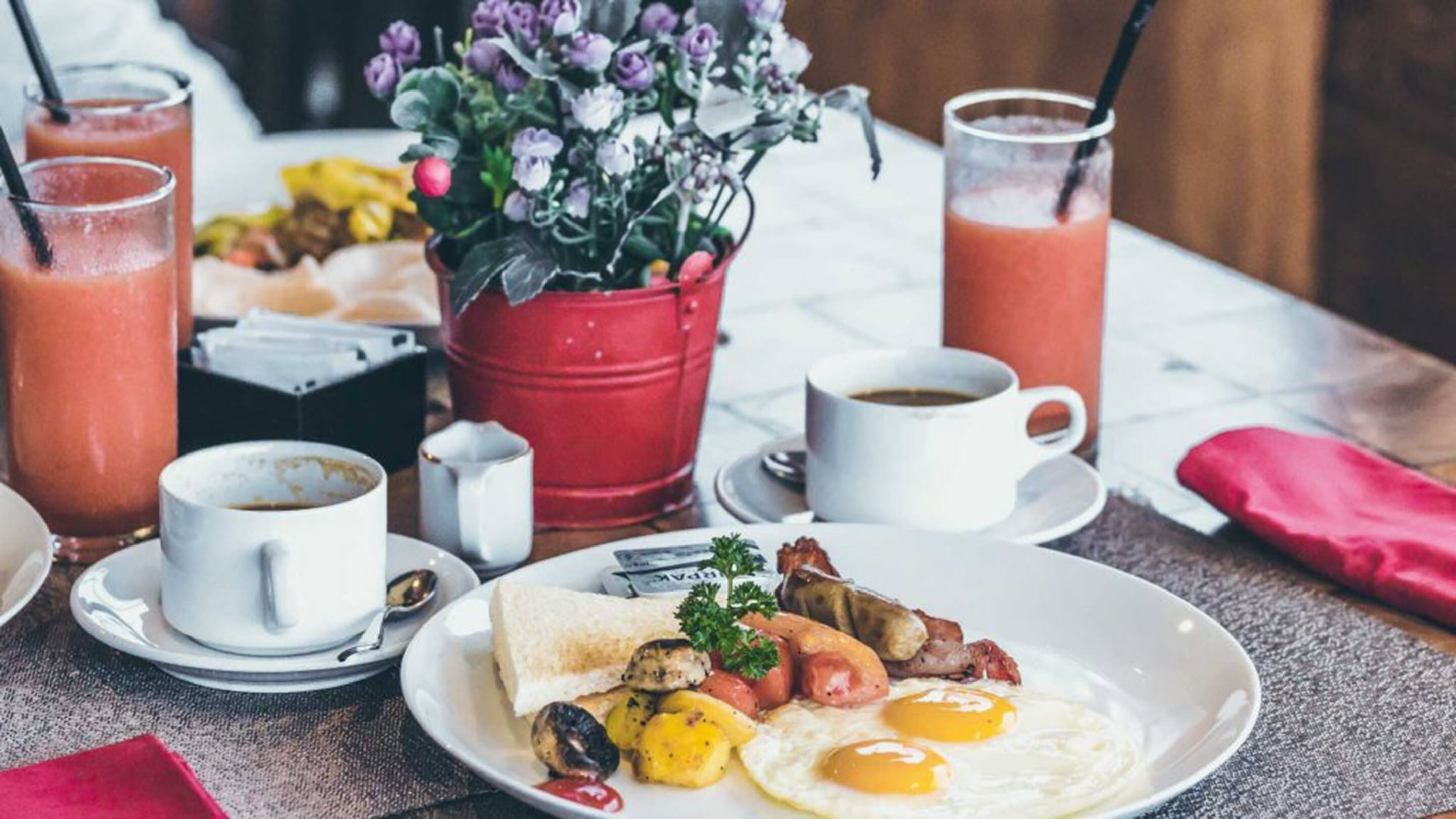 Mesa de café da manhã com prato de ovos, linguiça e torradas, dois copos de uma bebida vermelha, uma xícara de café e um balde vermelho com flores.