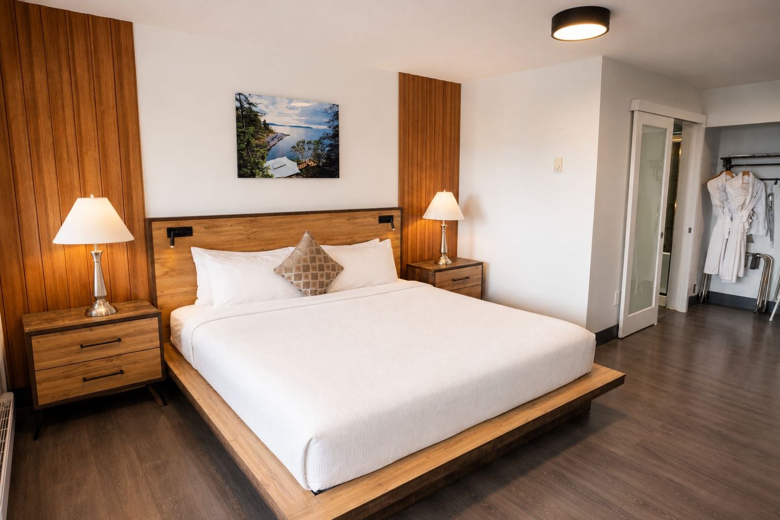 Quarto de hotel com uma cama grande, cabeceira de madeira, duas luminárias laterais, um quadro na parede e um porta-robes.