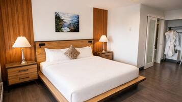 Quarto de hotel com uma cama grande, cabeceira de madeira, duas luminárias laterais, um quadro na parede e um porta-robes.