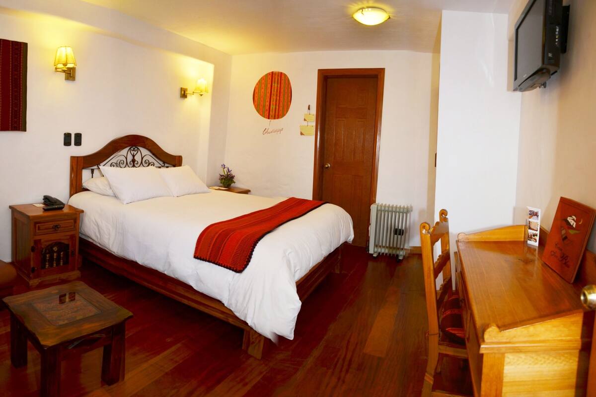Quarto de hotel bem arrumado, com uma cama de madeira, mesas de cabeceira, uma mesinha, uma cadeira e um radiador.