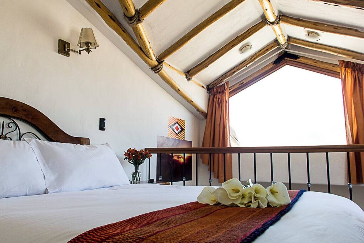 Quarto de hotel com cama, janela com cortinas, televisão fixada na parede e um vaso decorativo com flores.