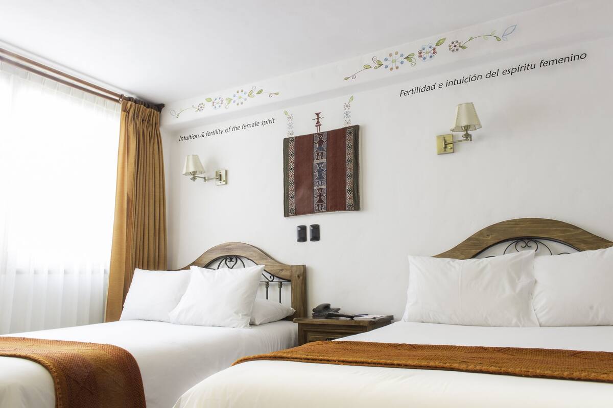 Quarto de hotel com duas camas, roupa de cama branca, cabeceira de madeira, parede decorativa com uma citação e uma janela com cortinas.