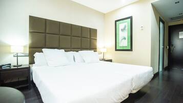 Quarto de hotel com uma cama grande, duas mesas de cabeceira com abajures, uma obra de arte emoldurada na parede e uma porta à direita.