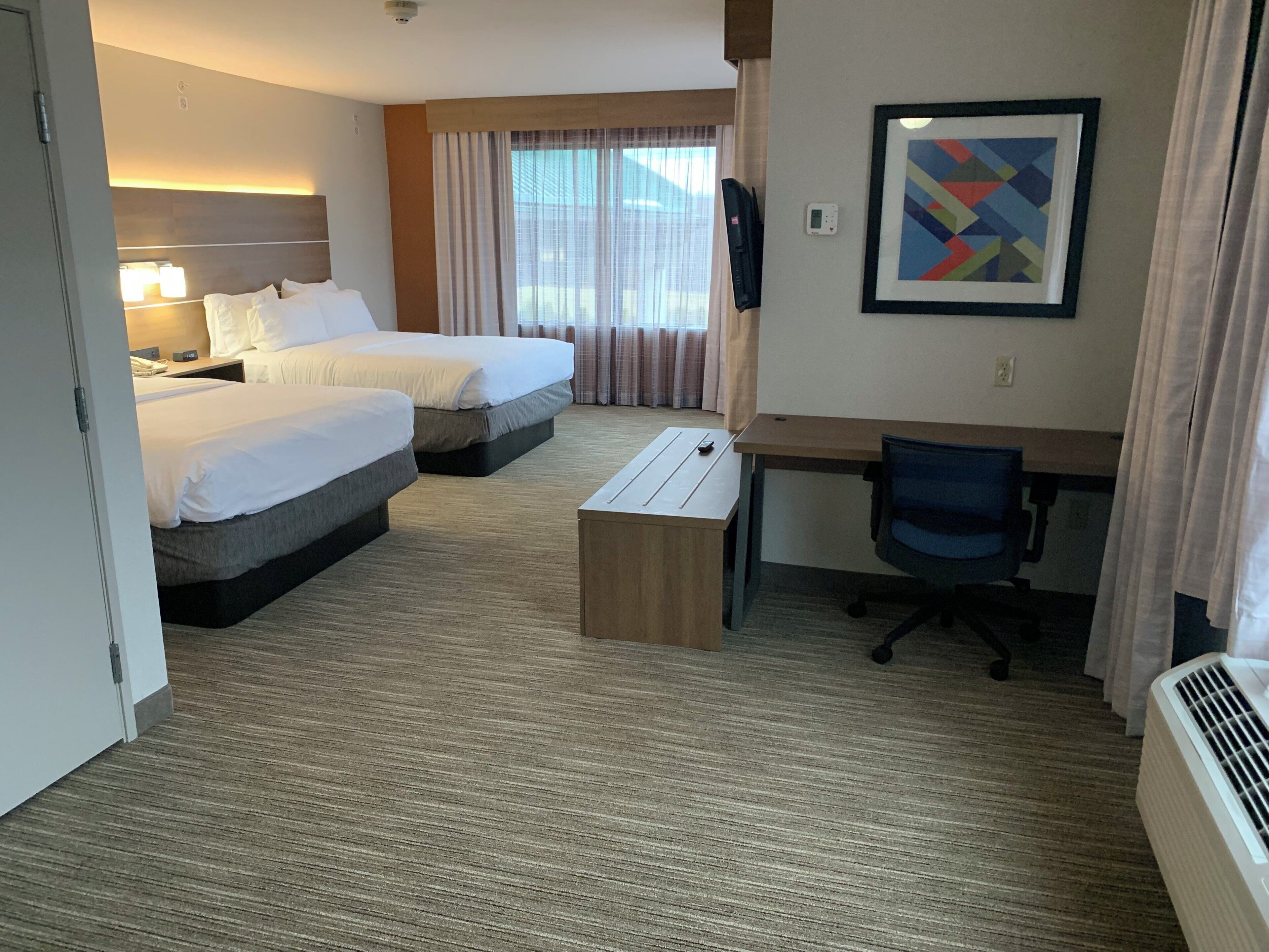 Quarto de hotel com duas camas, uma escrivaninha, uma cadeira, uma mesinha, um aquecedor, uma televisão e uma janela com cortinas.