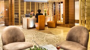 Um saguão de hotel com uma mesa de recepção, duas poltronas, uma mesa de vidro com um laptop, uma bebida e uma bandeja de comida.