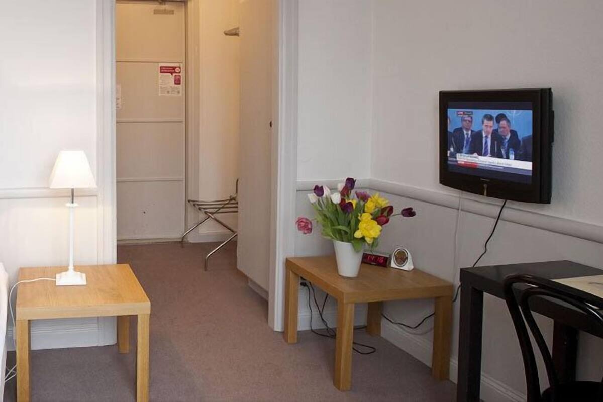 Quarto de hotel com cama, escrivaninha, cadeira, televisão e uma pequena mesa com um vaso de flores.