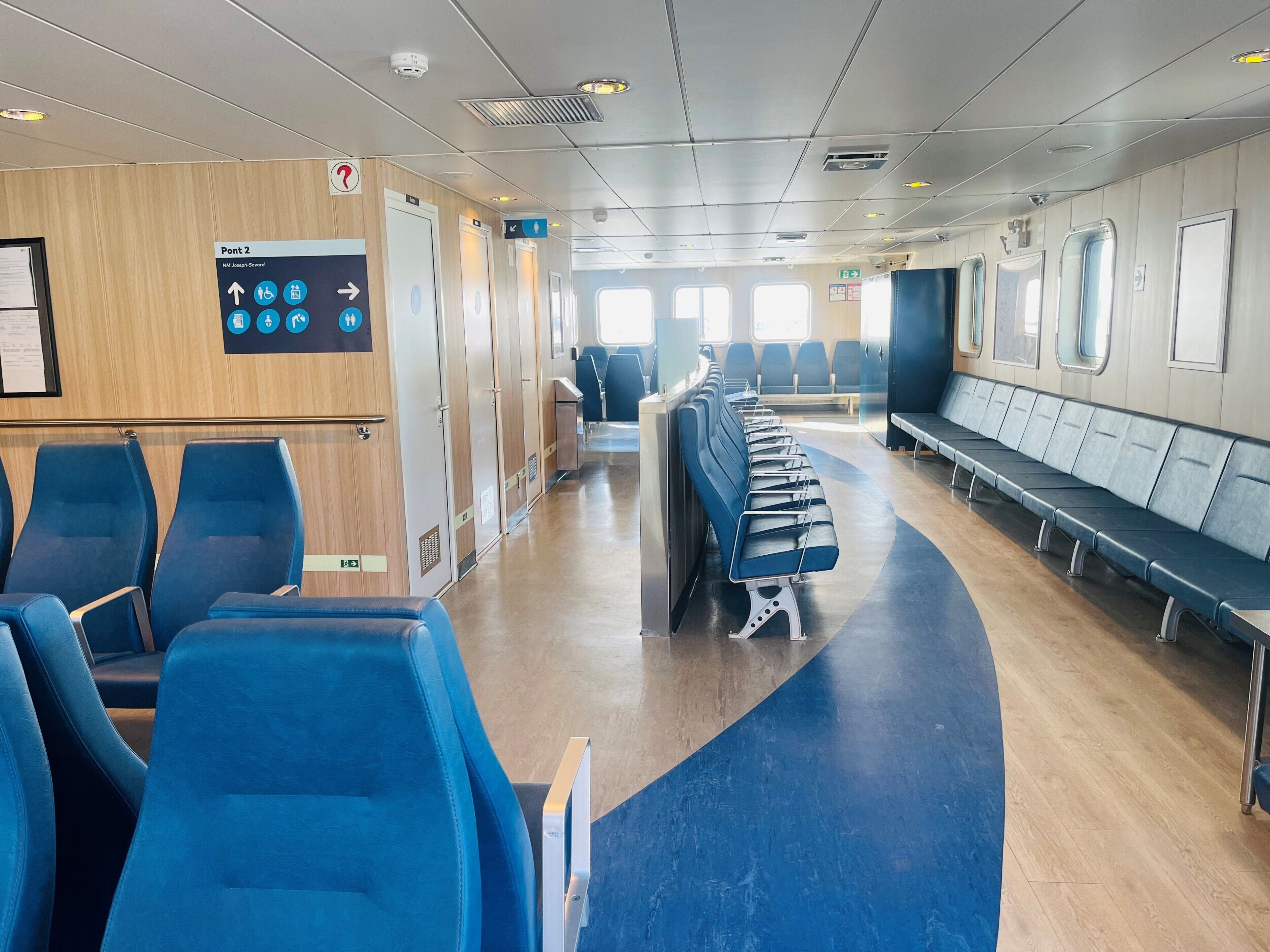 Interior de um vagão de trem com assentos azuis, painel de parede de madeira e uma placa indicando 'Pont 2 NM Joseph-Savard'.