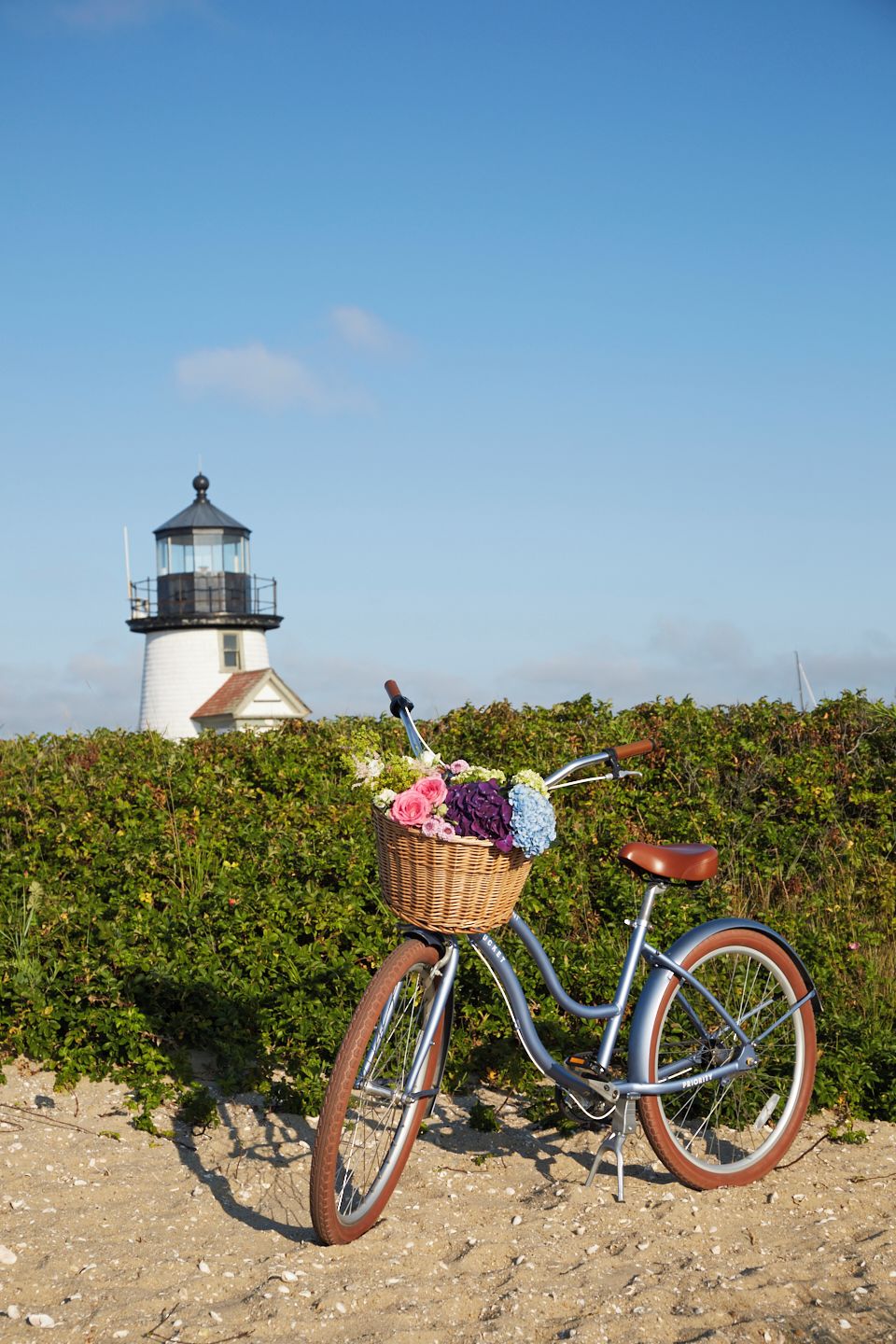 Uma bicicleta com um cesto cheio de flores, estacionada em um caminho de cascalho, com um farol ao fundo.