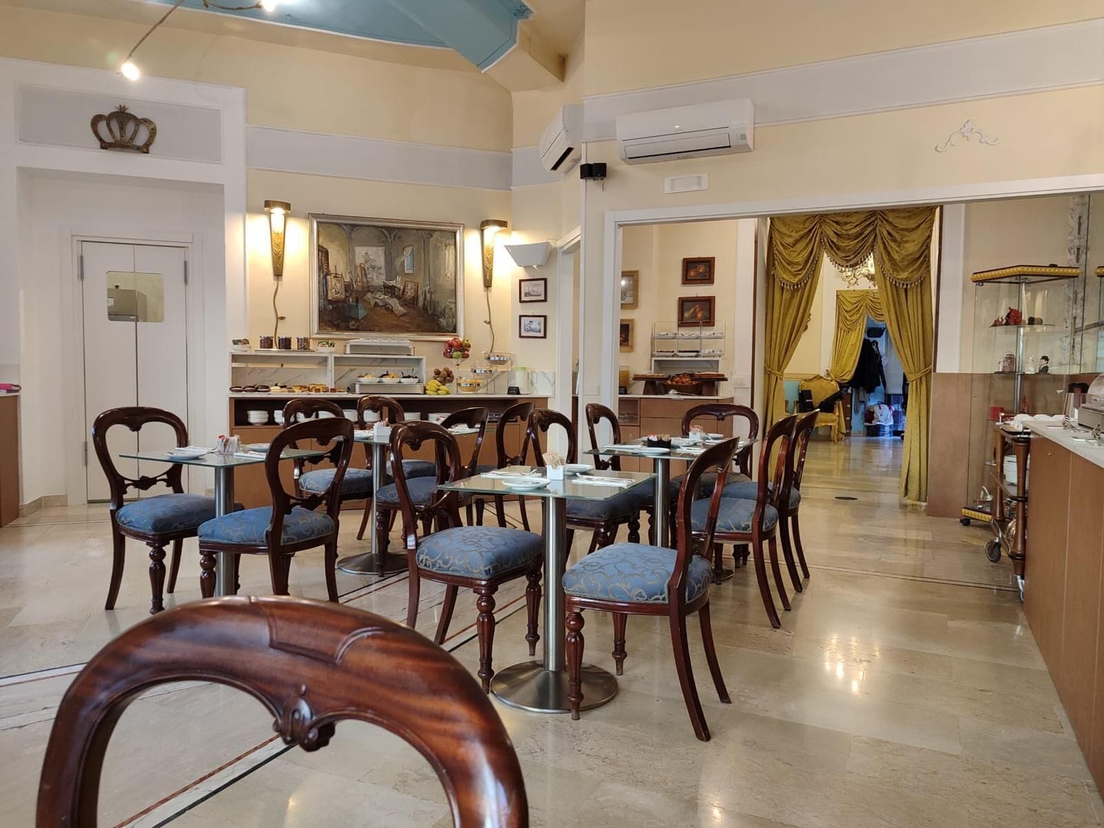 Uma sala de jantar com cadeiras e mesas de madeira, uma cozinha ao fundo e uma porta de acesso a outro espaço.