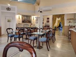 Uma sala de jantar com cadeiras e mesas de madeira, uma cozinha ao fundo e uma porta de acesso a outro espaço.
