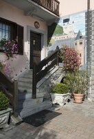 Uma escadaria de pedra leva a um edifício com guarda-corpo de madeira escura e uma porta. Há plantas em vasos com flores cor-de-rosa nos degraus.