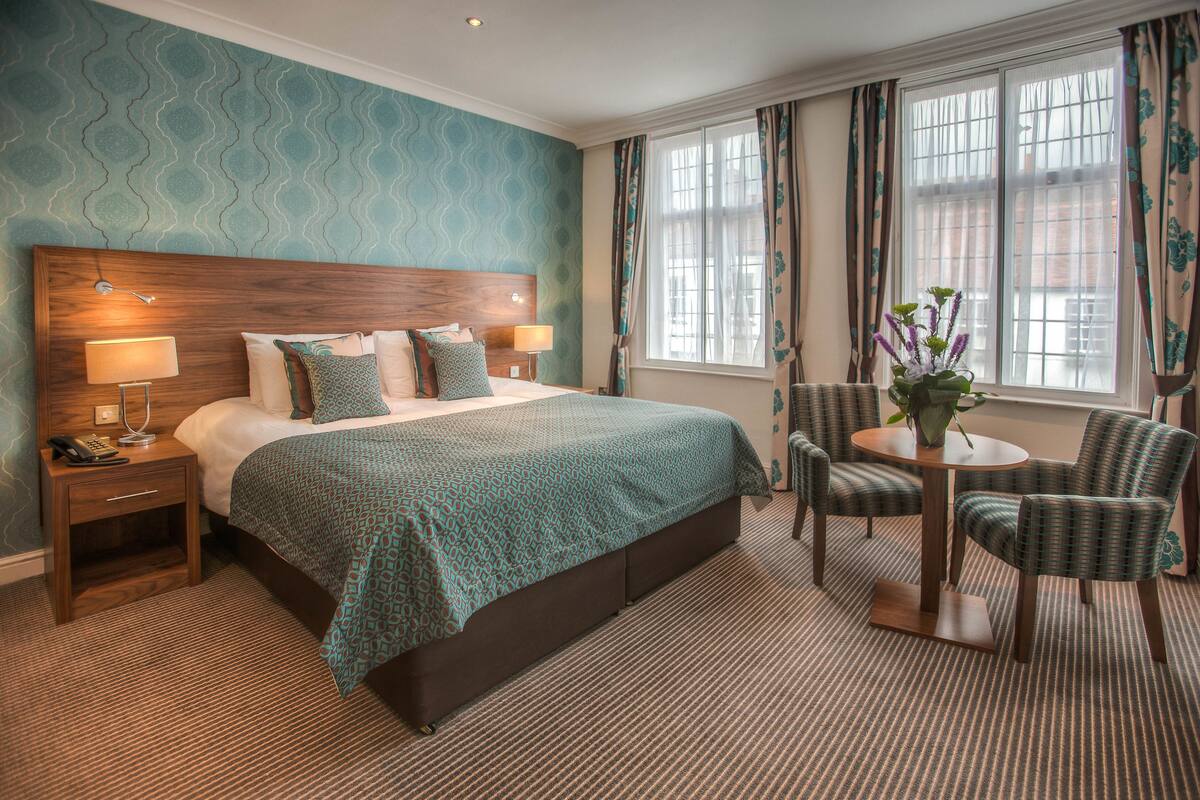 Quarto de hotel com uma cama grande, mesa de cabeceira com telefone, uma cadeira, mesa com um vaso e janela com cortinas.