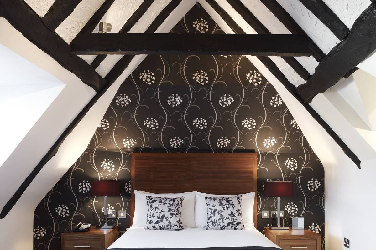 Quarto com teto inclinado, cabeceira de madeira e papel de parede estampado. Há duas mesas de cabeceira com luminárias, uma cama com edredom preto e branco, e piso coberto por carpete.