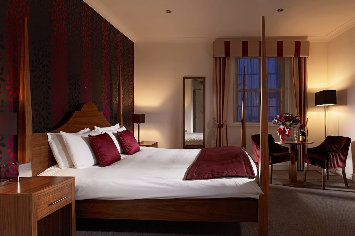Quarto de hotel com cama grande, mesa de cabeceira, cadeira, pequena mesa com um vaso de flores e janela com cortinas.