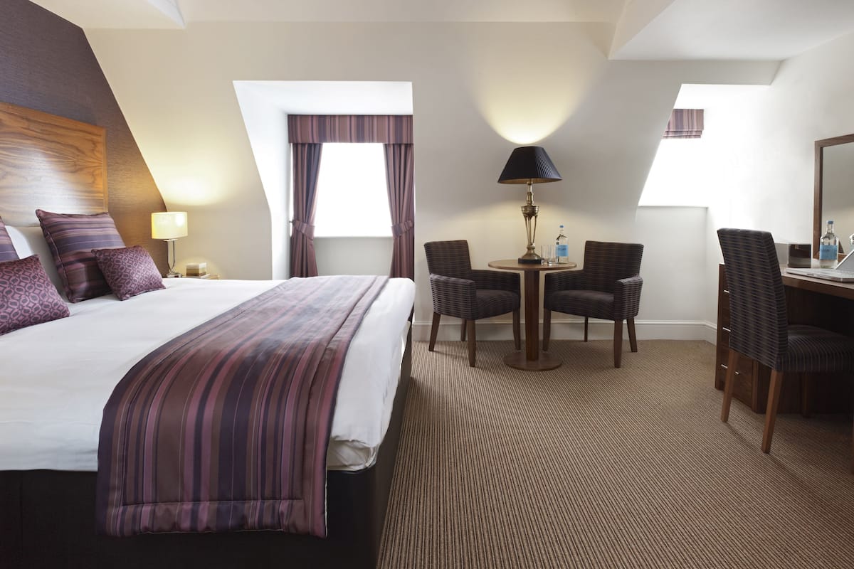Quarto de hotel com uma cama grande, duas cadeiras, uma mesa pequena e uma escrivaninha com um laptop.