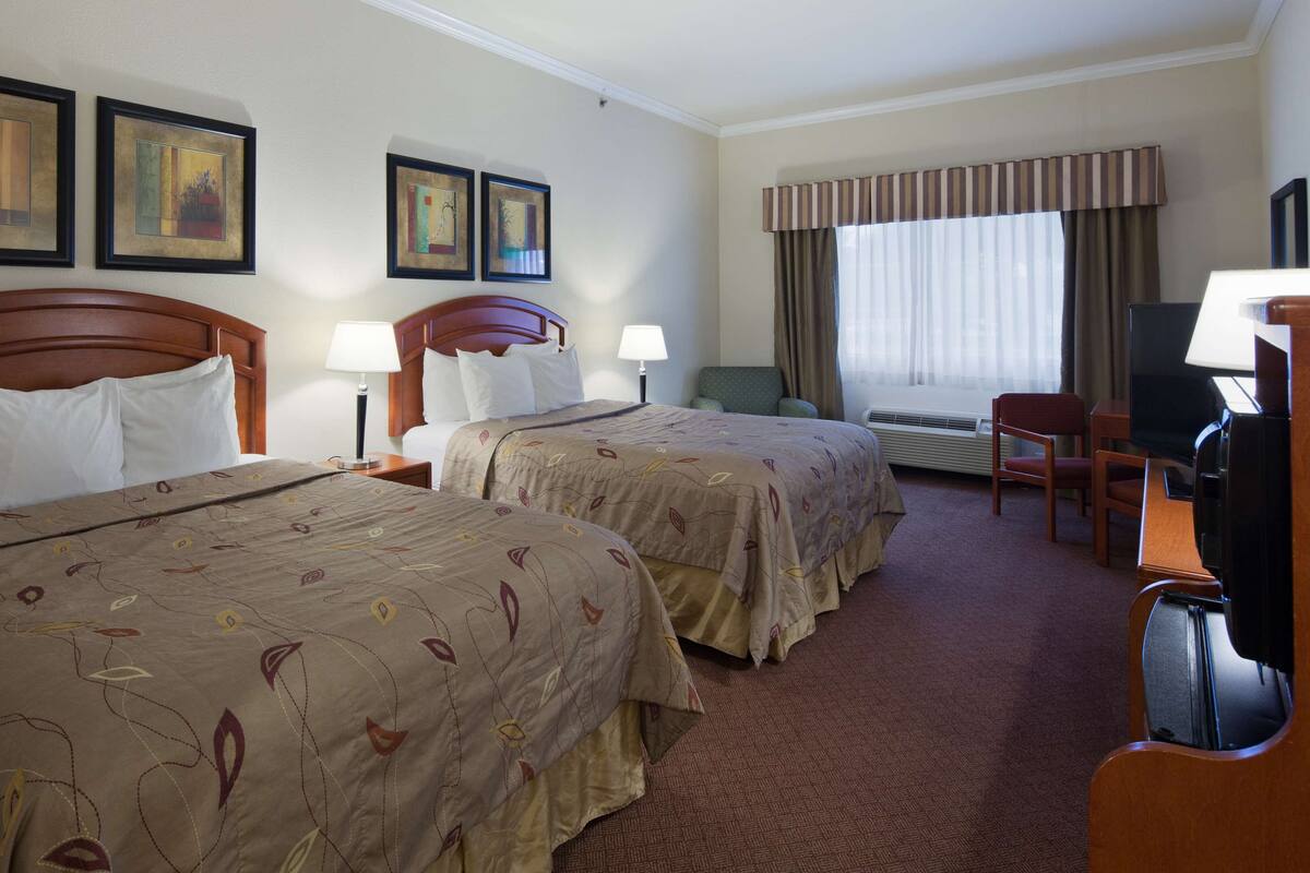 Quarto de hotel com duas camas, televisão, escrivaninha e uma cadeira. Há quadros nas paredes e um aparelho de ar condicionado.