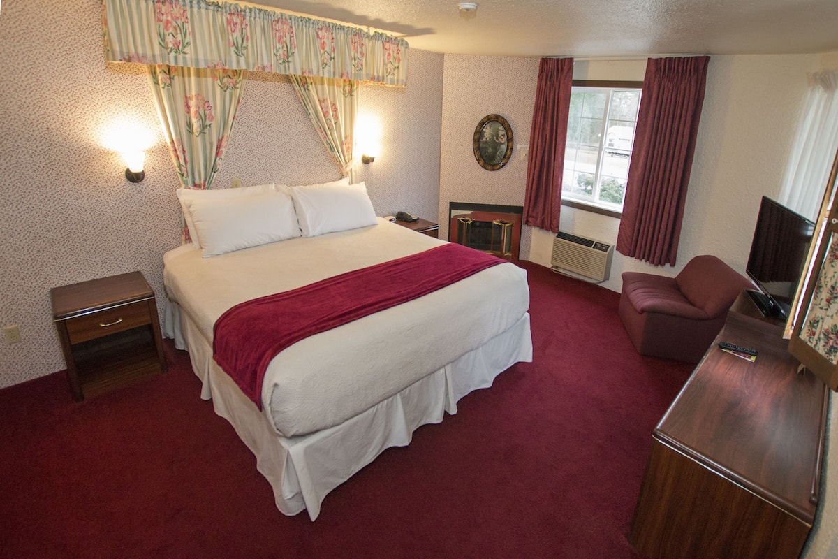 Quarto de hotel com uma cama grande, uma mesinha de cabeceira, uma cadeira, uma televisão e uma janela com cortinas.